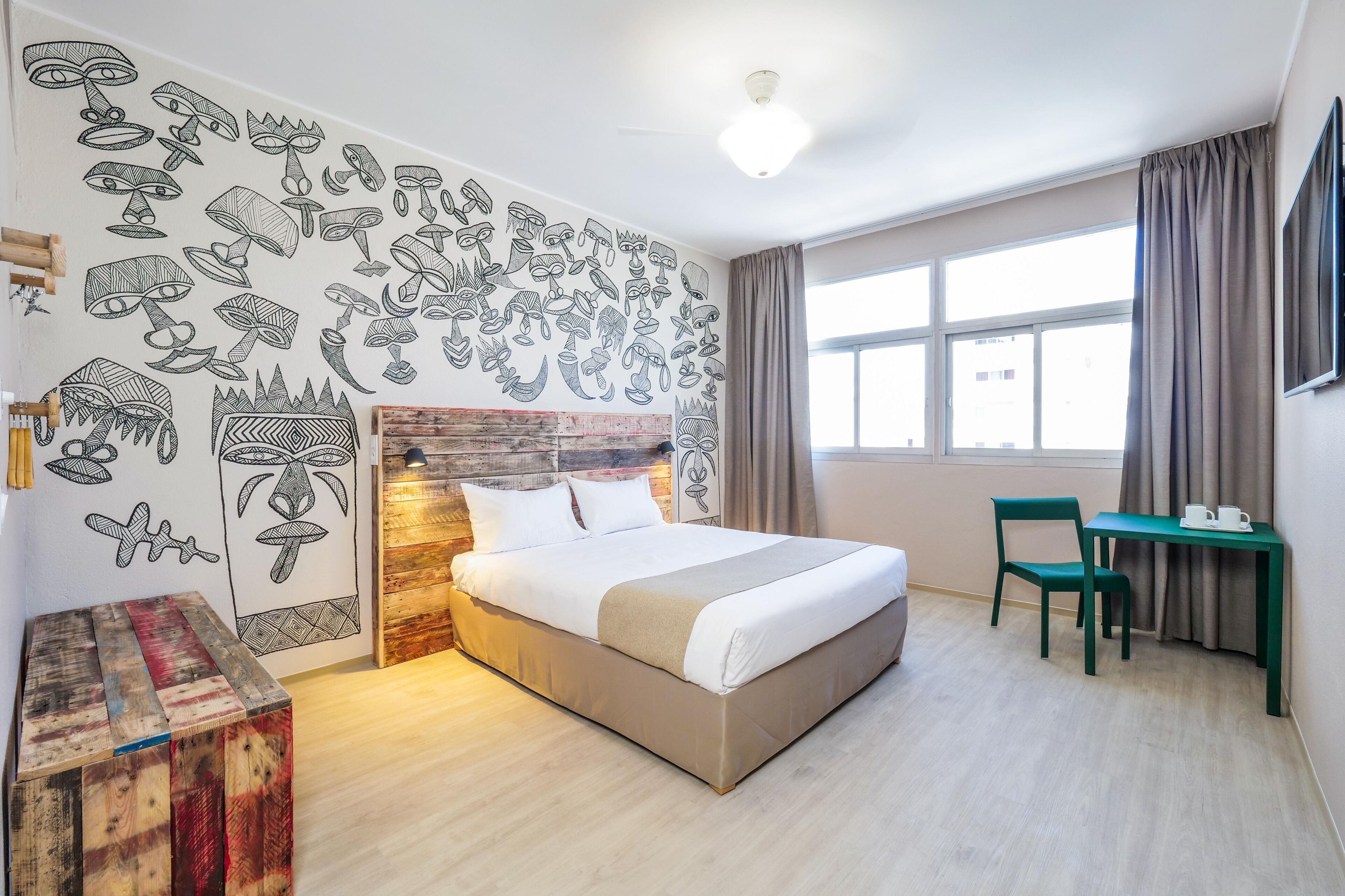 Quarto de hotel moderno com uma cama grande, cabeceira de madeira, escrivaninha com cadeira e janela com cortinas.