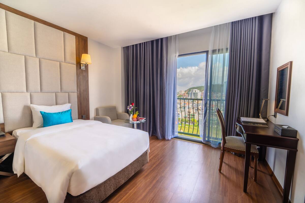Quarto de hotel com uma cama grande, uma escrivaninha, uma cadeira e vista para a cidade pela janela.