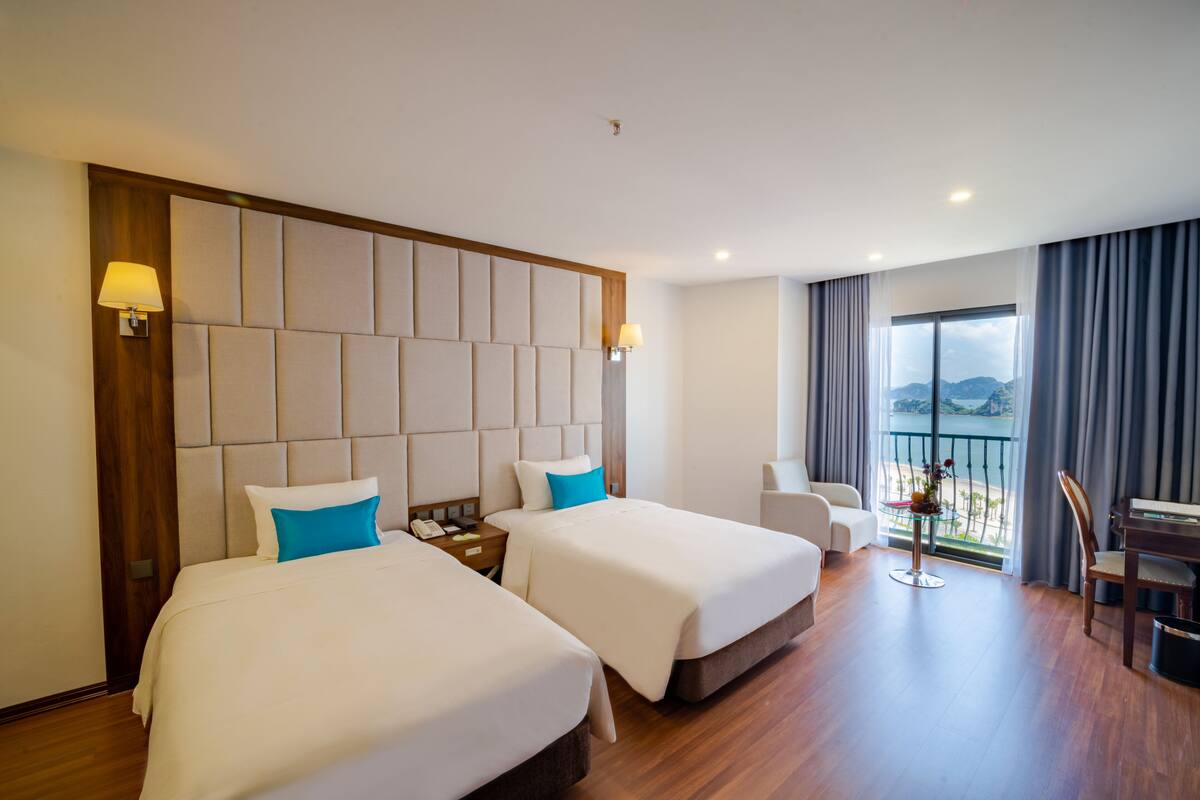 Quarto de hotel com duas camas, uma escrivaninha, uma cadeira, varanda com vista e cabeceira de madeira.
