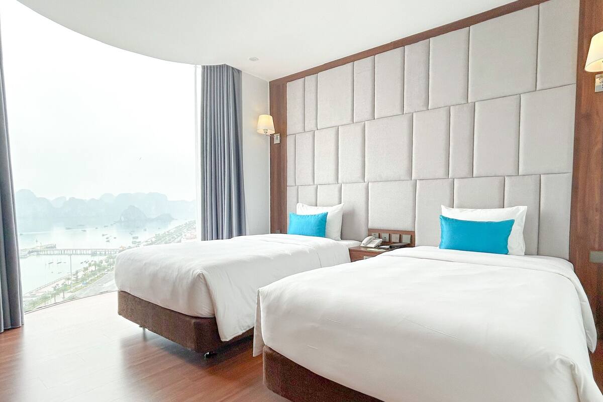 Quarto de hotel com duas camas, uma cabeceira grande e acolchoada, uma mesa de cabeceira com abajur e vista para a paisagem urbana através de grandes janelas.