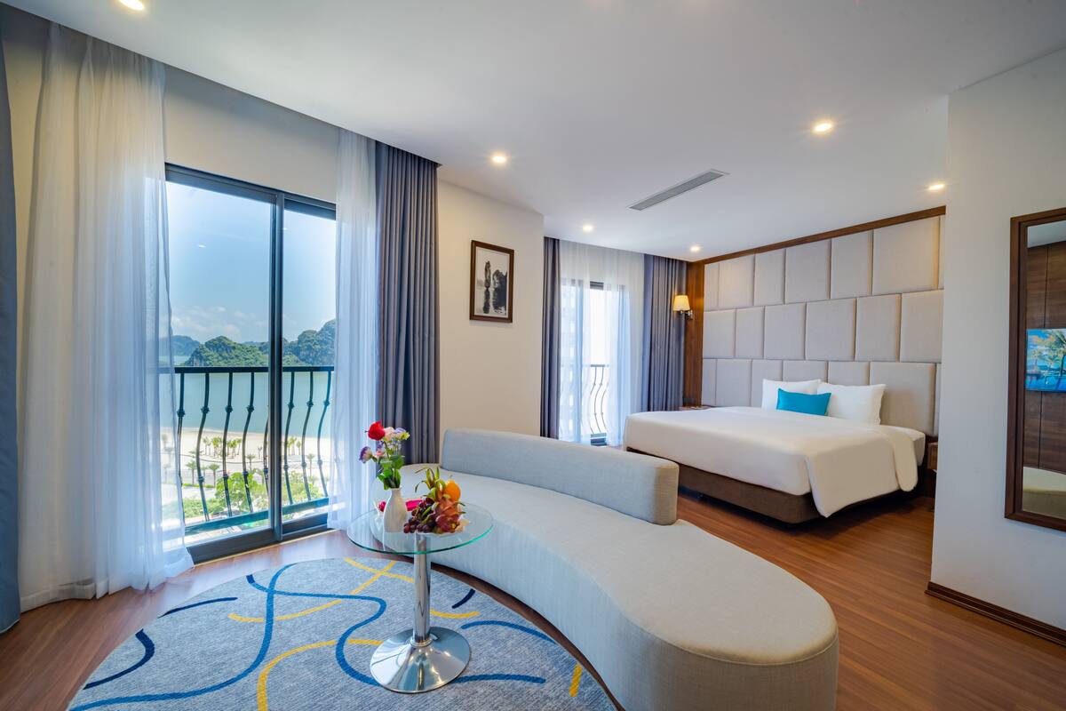Quarto de hotel moderno com uma cama grande, um sofá, uma mesa de vidro com flores e uma varanda com vista.