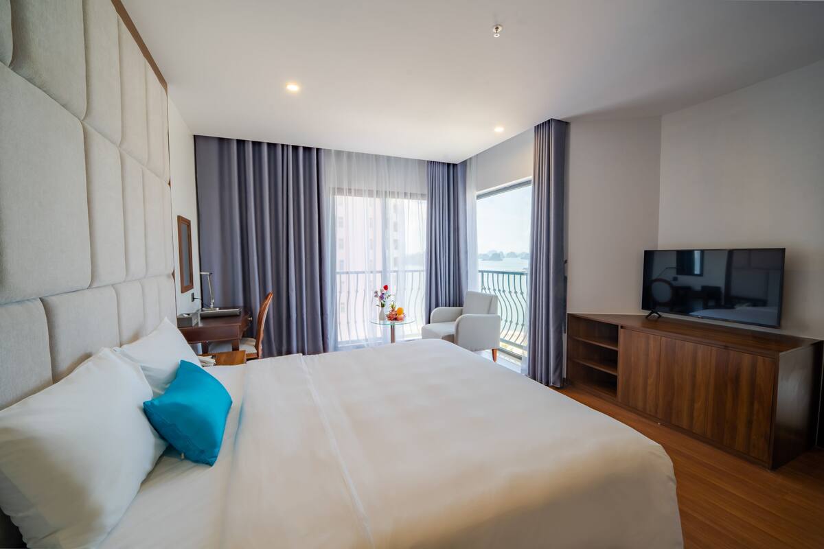 Quarto de hotel com uma cama grande, televisão, escrivaninha e varanda com cortinas.