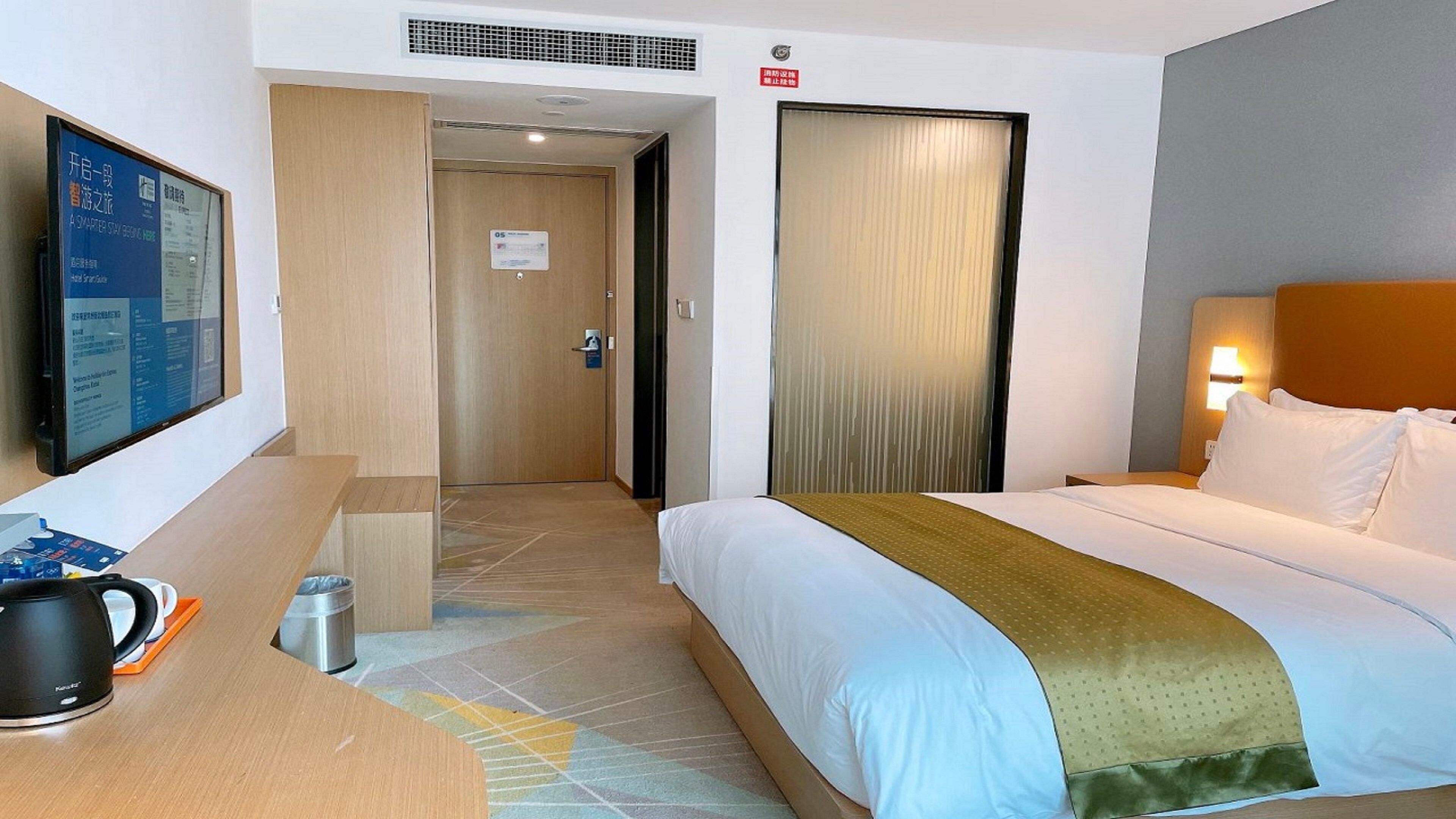 Quarto de hotel com cama, escrivaninha com cafeteira, televisão e porta que dá para outro quarto.