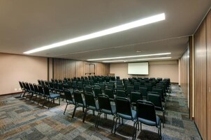 Uma sala de conferências com fileiras de cadeiras, uma tela de projetor e piso carpeteado.