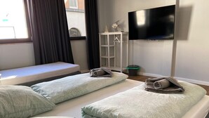 Quarto de hotel com três camas, uma TV fixada na parede, janela com cortinas e uma pequena estante com objetos decorativos.