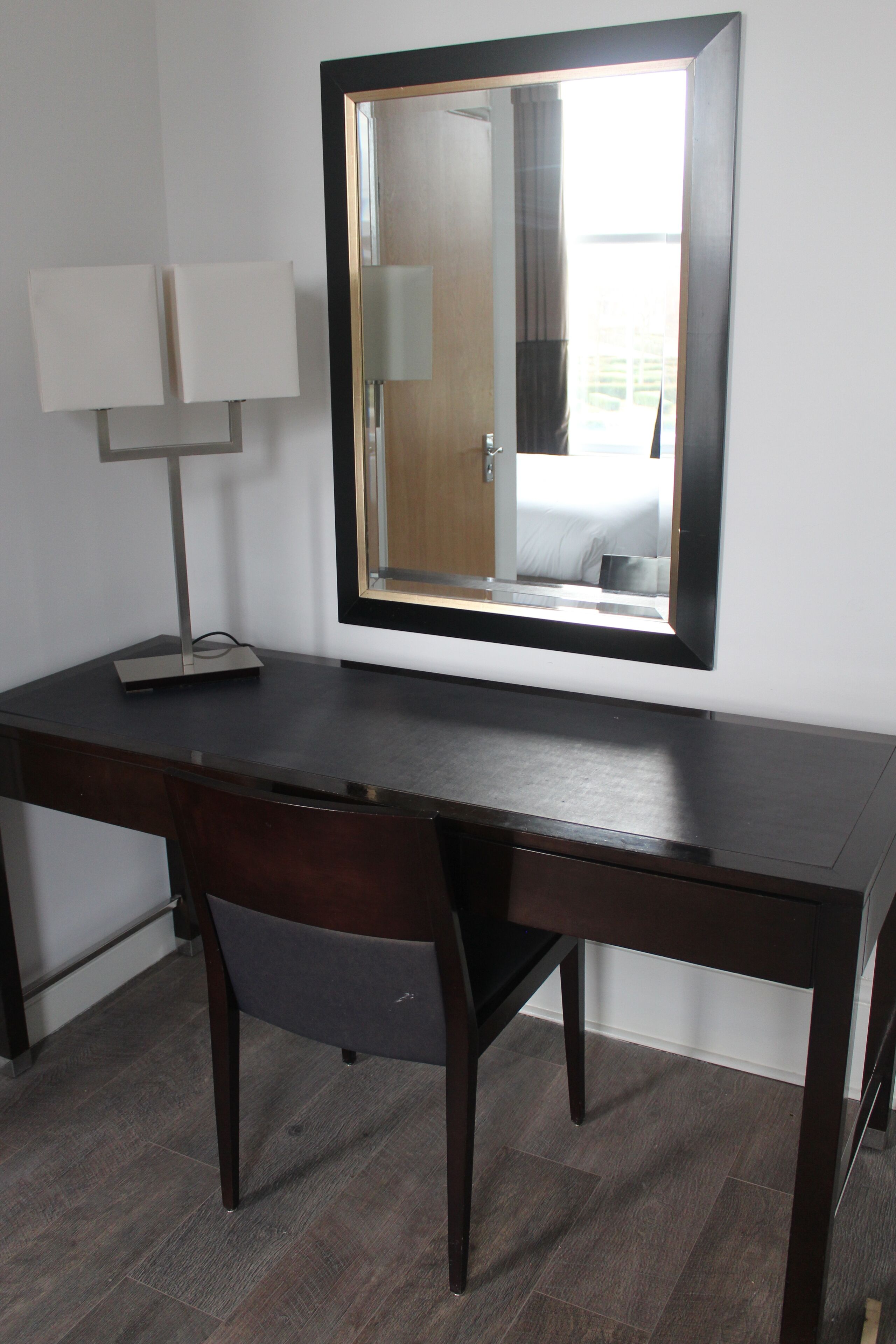 Uma mesa de madeira escura com uma luminária moderna, uma cadeira e um espelho grande que reflete um quarto com cama e cortinas.