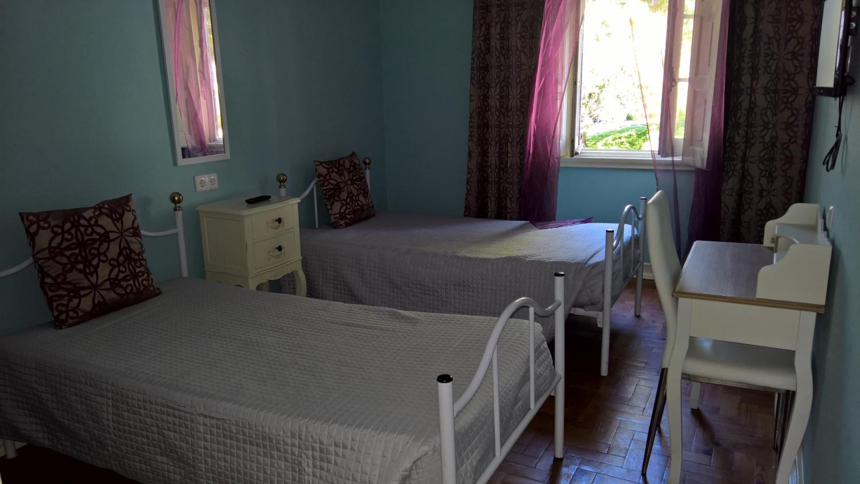 Quarto com duas camas, uma mesa de cabeceira, uma cadeira e janela com cortinas.