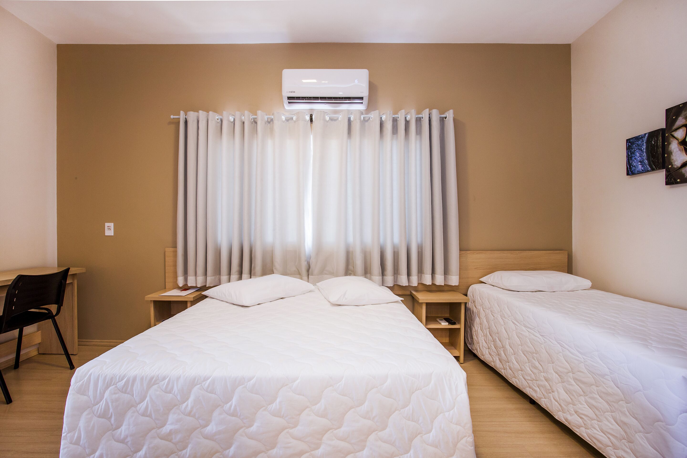 Quarto de hotel com uma cama grande, duas mesas de cabeceira, uma escrivaninha e um ar condicionado.