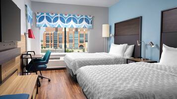 Quarto de hotel com duas camas, uma escrivaninha, uma cadeira e uma janela grande com vista para a cidade.