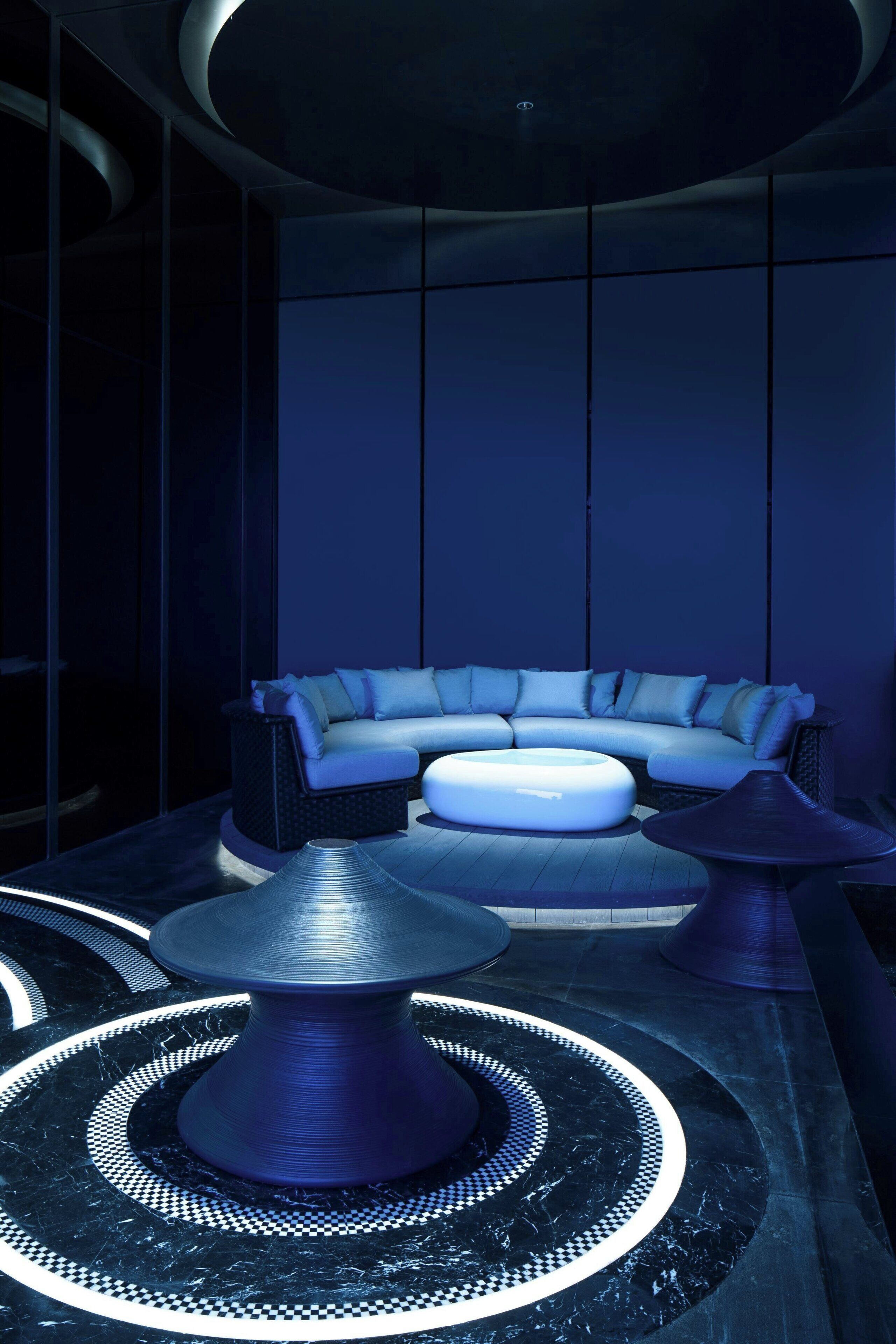 Uma área de estar moderna com um sofá curvo, um banquinho redondo e uma mesa central com design de iluminação circular.