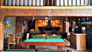 Uma sala de bilhar com duas mesas verdes, uma parede de tijolos e um espaço com balcão de madeira.
