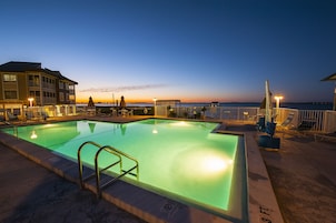 Piscina com vista para o pôr do sol, cadeiras de lounge e um edifício com vários andares.