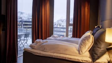 Quarto de hotel com cama, cadeira, mesa e vista para uma montanha nevada e edifícios.