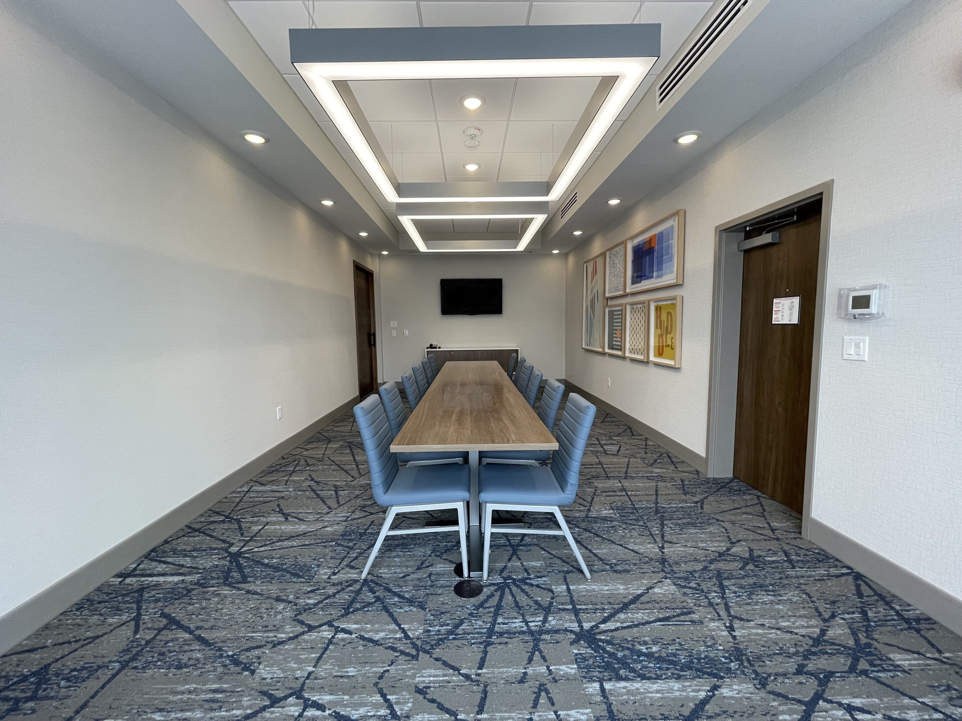 Sala de conferências com uma mesa comprida, cadeiras azuis, televisão de tela plana e carpete estampado.