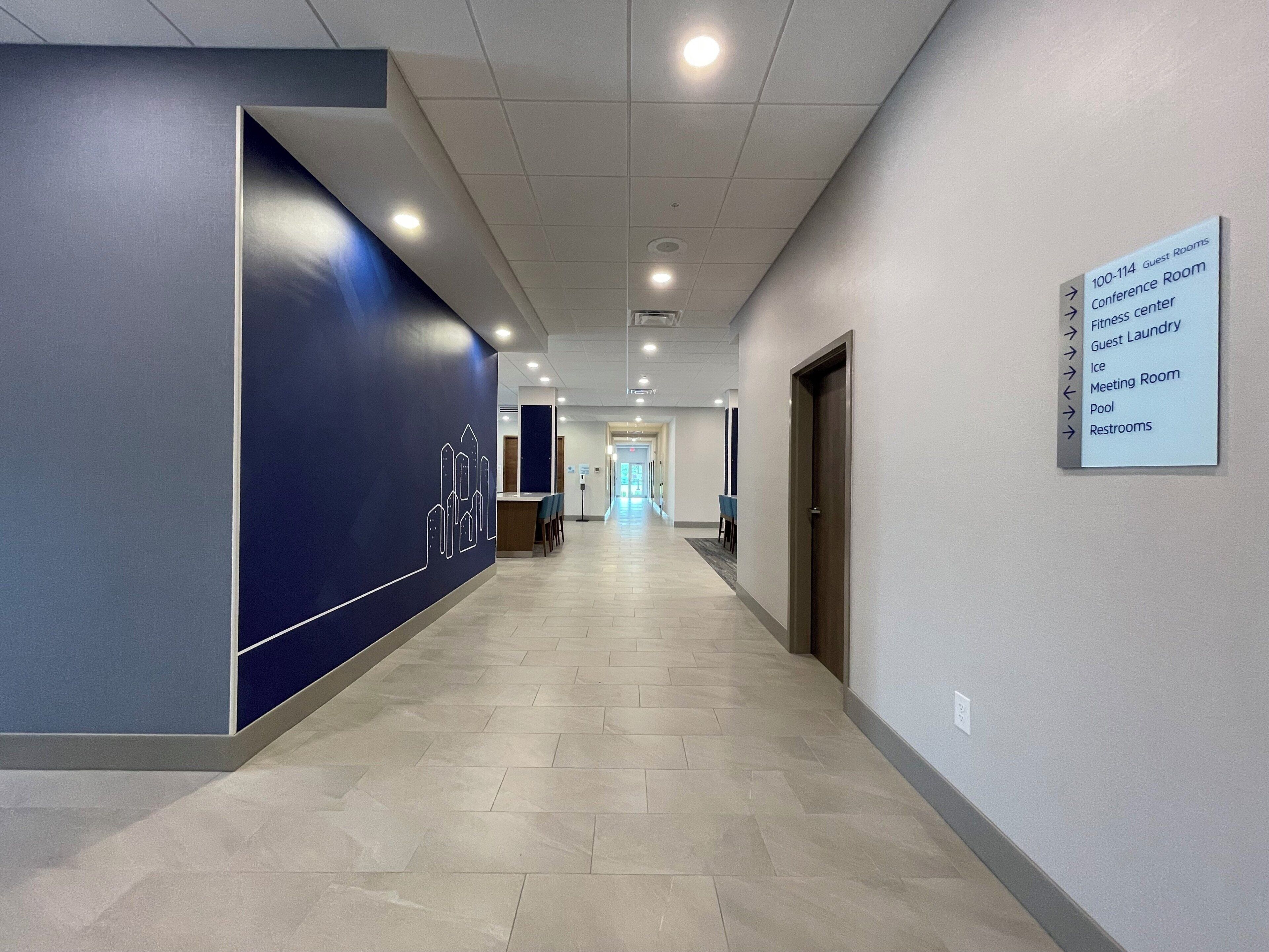 Corredor de hotel com aviso listando comodidades como quartos de hóspedes, sala de conferências, centro de atividades físicas, lavanderia para hóspedes, máquina de gelo, sala de reuniões, piscina e sanitários.