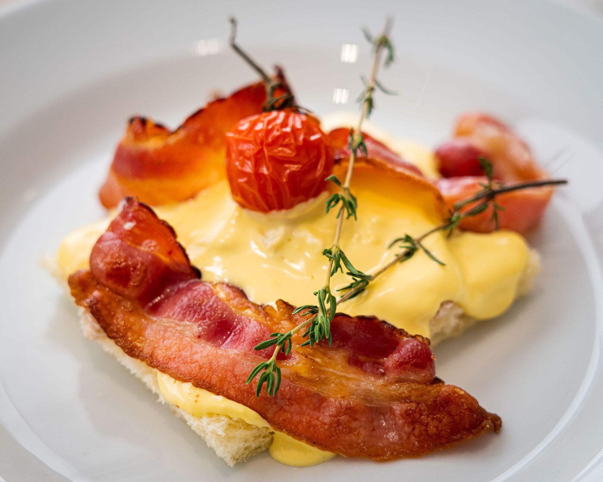 Um prato com uma fatia de torrada coberta com queijo derretido, uma fatia de bacon e um tomate cereja.