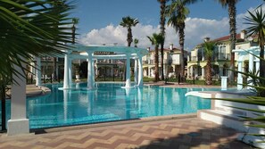 Piscina com pérgola branca, palmeiras e edifícios ao fundo.