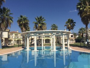 Uma piscina com uma estrutura circular branca no centro, rodeada por palmeiras e um edifício com varandas.