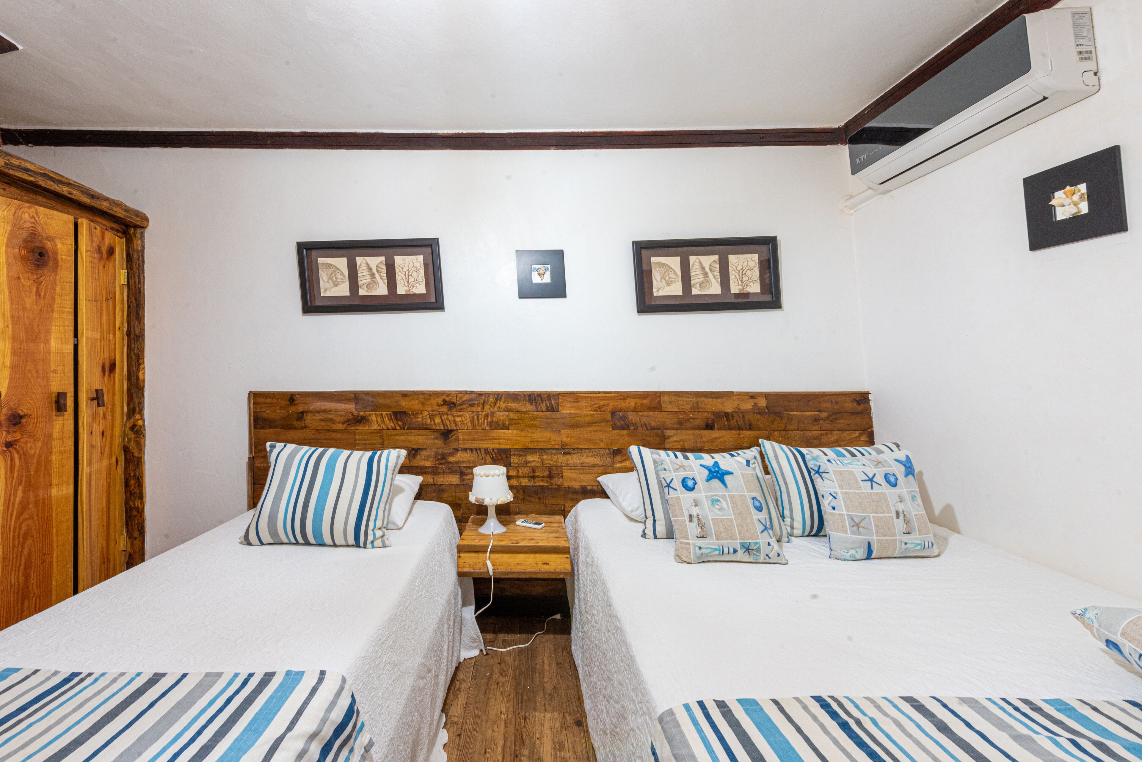 Quarto com duas camas, cabeceira de madeira, paredes brancas, quadros emoldurados e guarda-roupa de madeira.