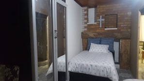 Quarto com cama, cabeceira de madeira, parede de tijolos e cruz decorativa.