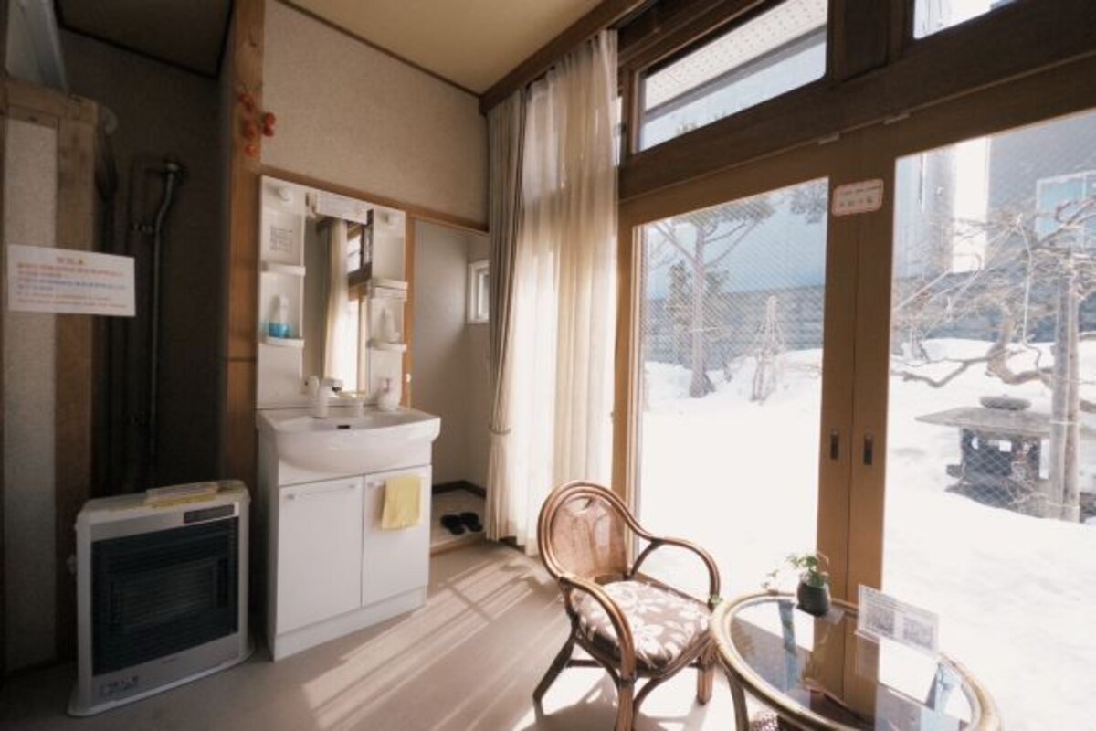 Quarto de hotel com um armário de madeira, uma mesinha, uma cadeira, um aquecedor e uma janela com vista para uma área externa coberta de neve.