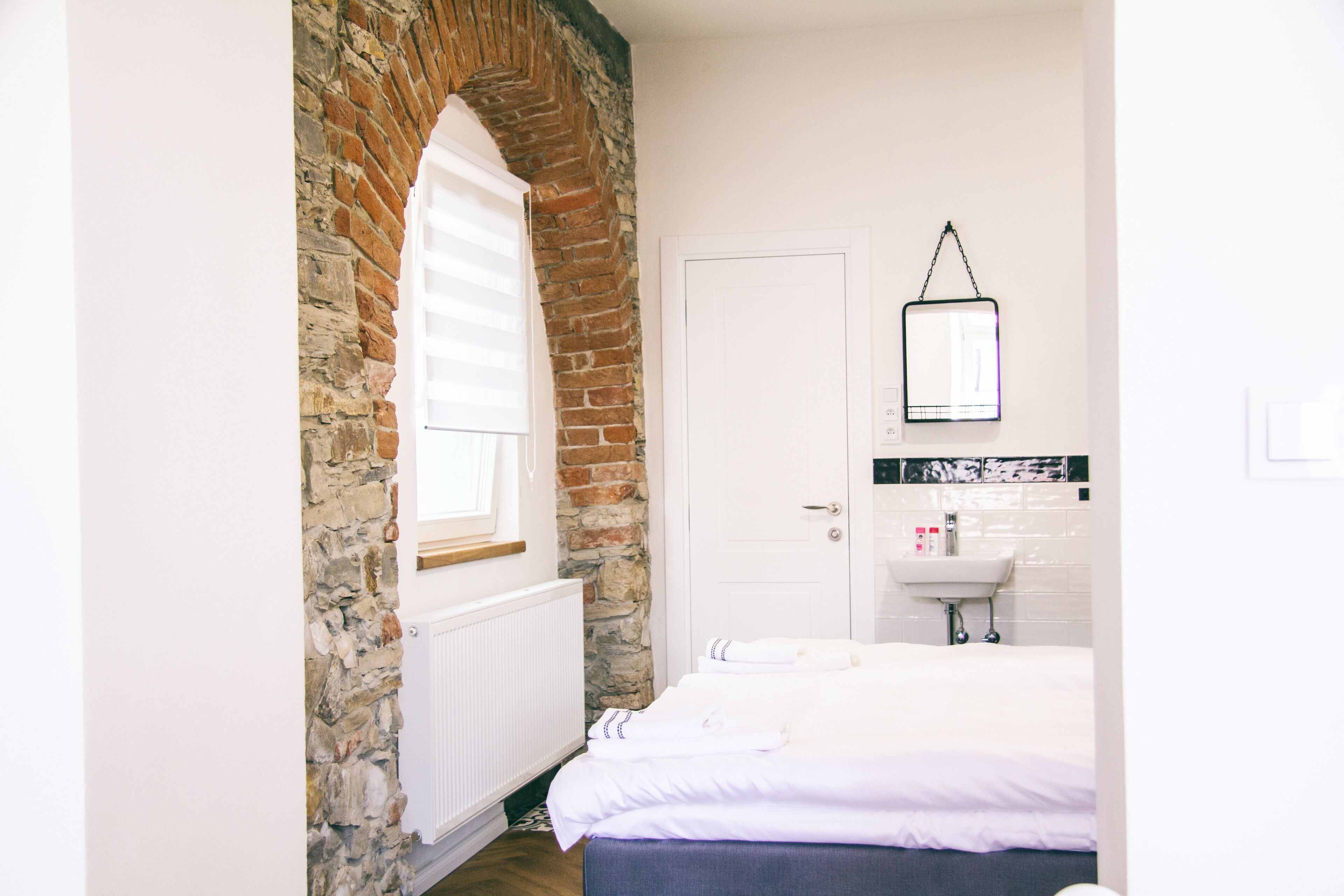 Quarto com arco de pedra, porta branca, cama com roupa de cama branca, luminária pendente e pia branca com painel de fundo preto.