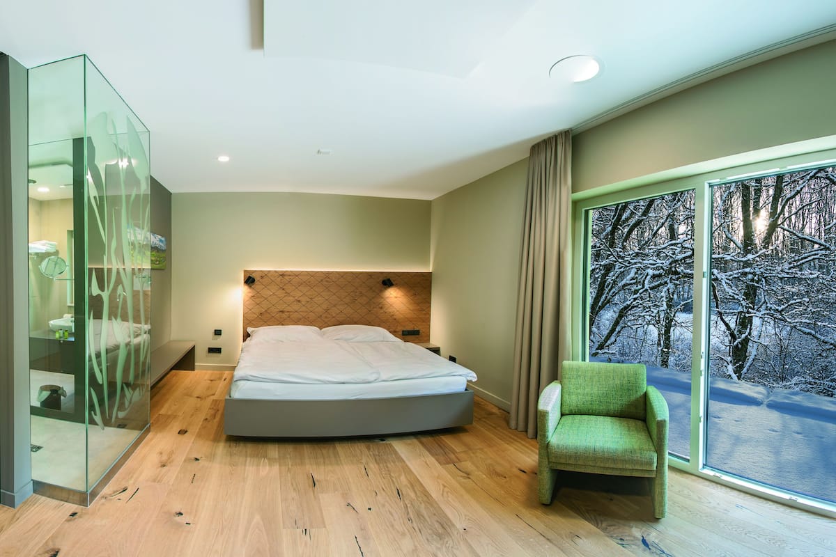 Quarto moderno com cama grande, divisória de vidro, cadeira verde e vista para uma floresta nevada através de uma janela ampla.