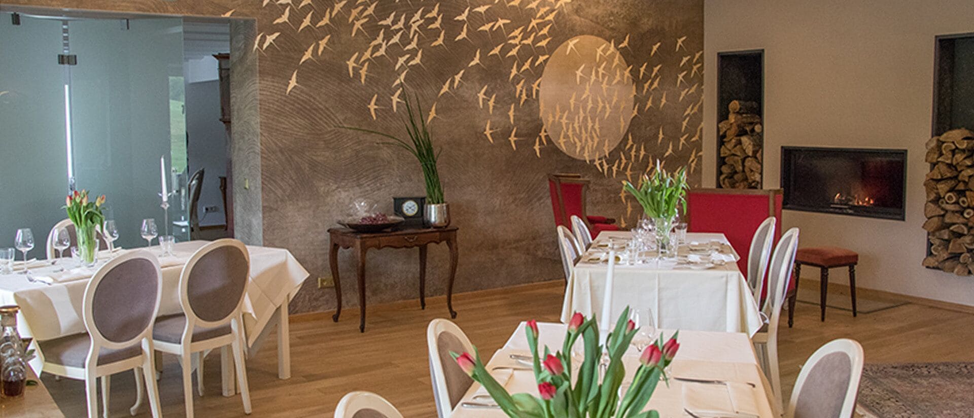 Uma sala de jantar com mesas preparadas para refeição, uma parede com mural de pássaros, lareira e um relógio em uma mesinha.