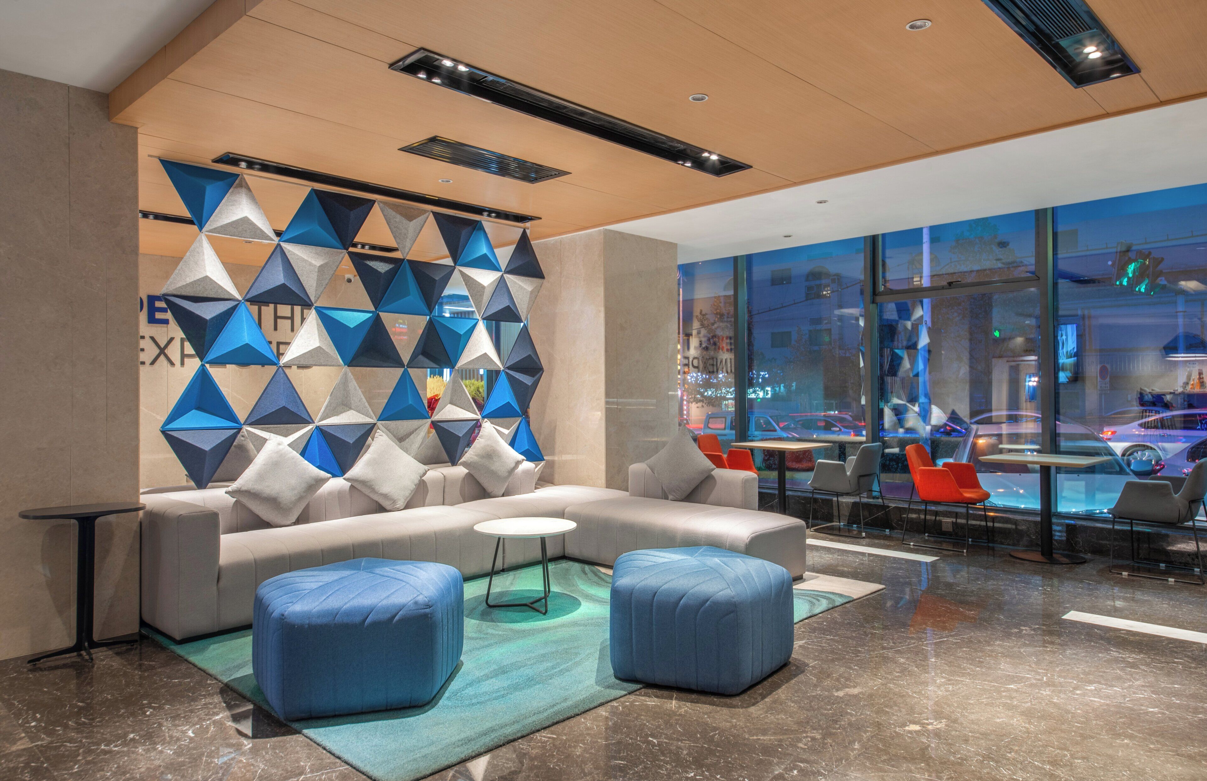 Um lobby moderno com design de parede geométrico, área de estar com sofá e pufes, e vista da cidade através de grandes janelas.