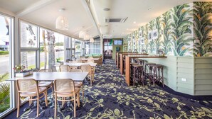 Um restaurante com grandes janelas, cadeiras de vime, um tapete estampado e um papel de parede tropical.