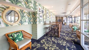 Um saguão de hotel com parede estampada com padrão de folhagem tropical, um espelho redondo, móveis de vime e um tapete com desenho de folhas.