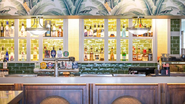 Uma área de bar com um mural de folhagem tropical, prateleiras cheias de garrafas de bebidas e um balcão com torneiras.