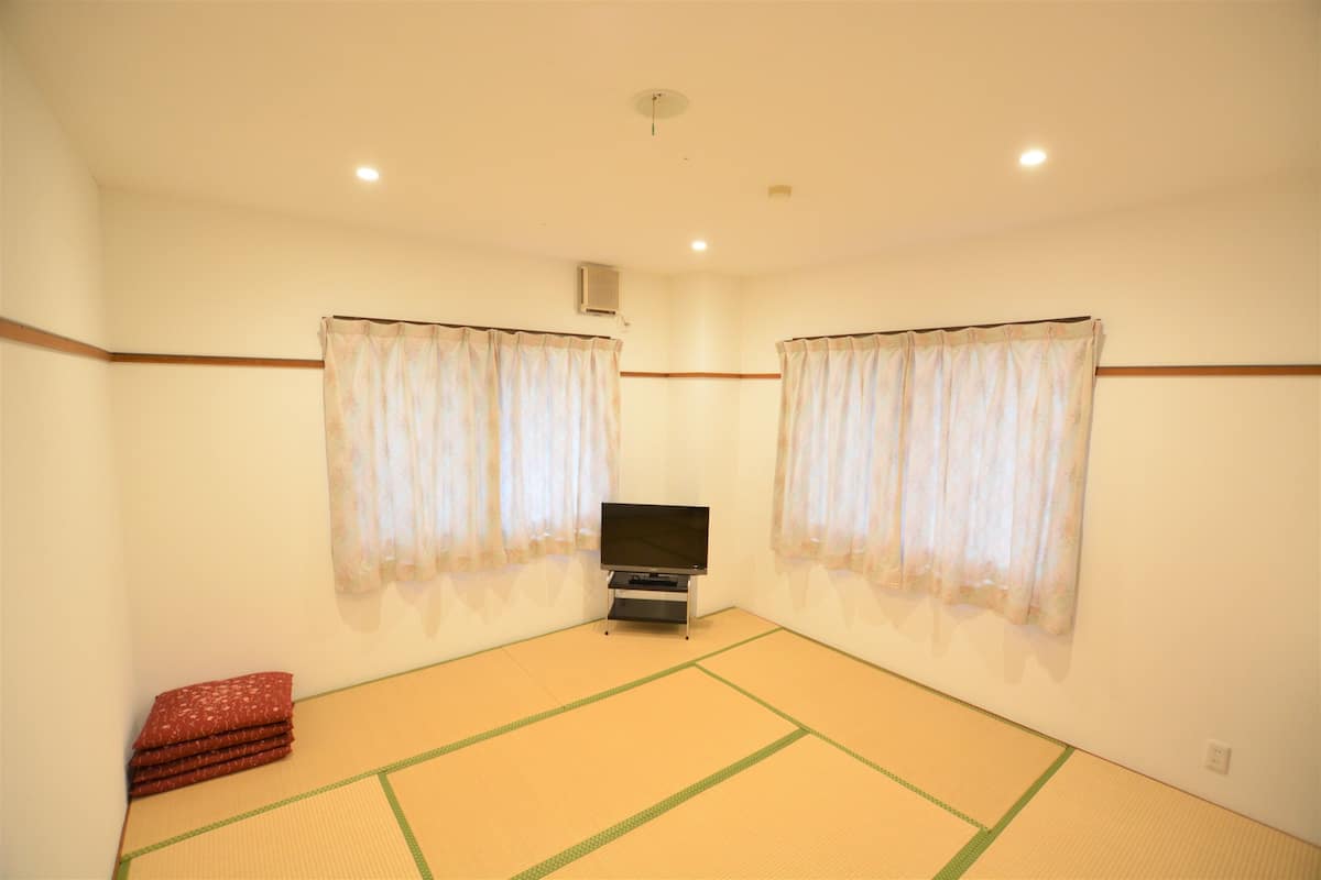 Quarto com tatames, uma televisão e duas janelas com cortinas.