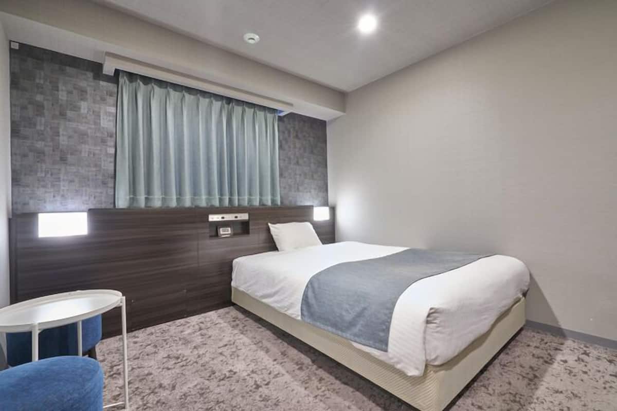 Quarto com cama, mesa de cabeceira, cadeira, janela com cortinas e luminária na parede.