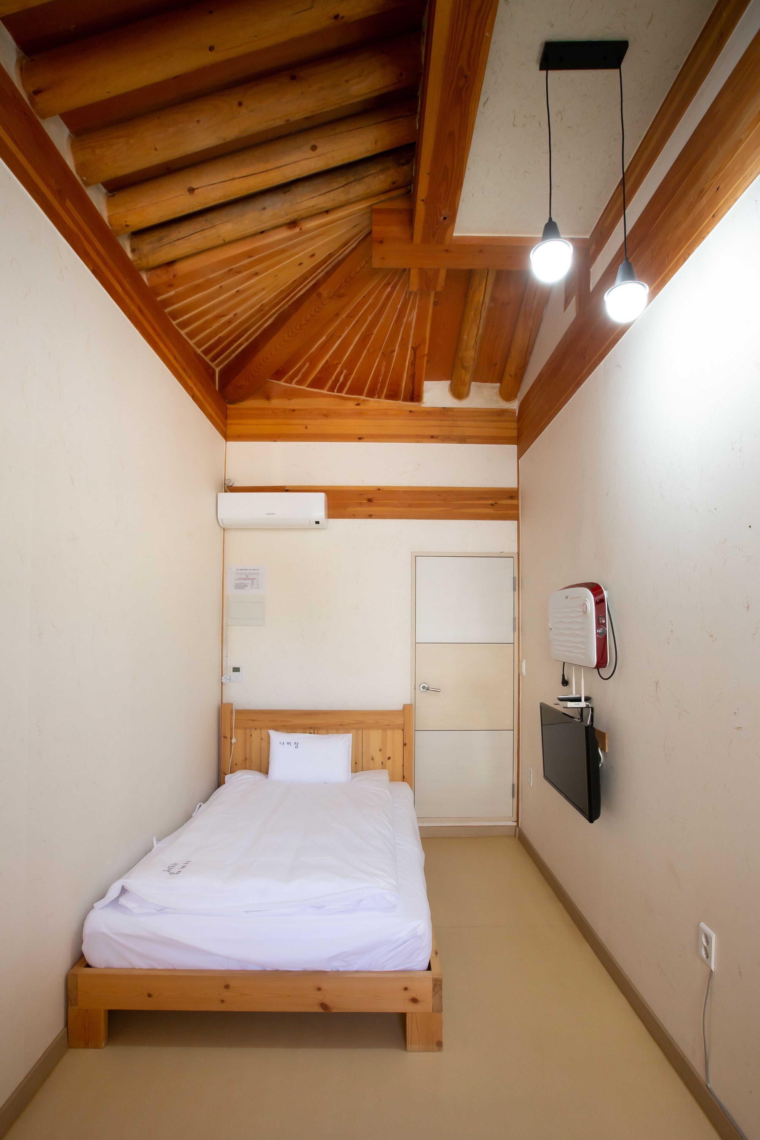 Um quarto pequeno e acolhedor, com uma cama de madeira, parede branca, ventoinha no teto e televisão montada.