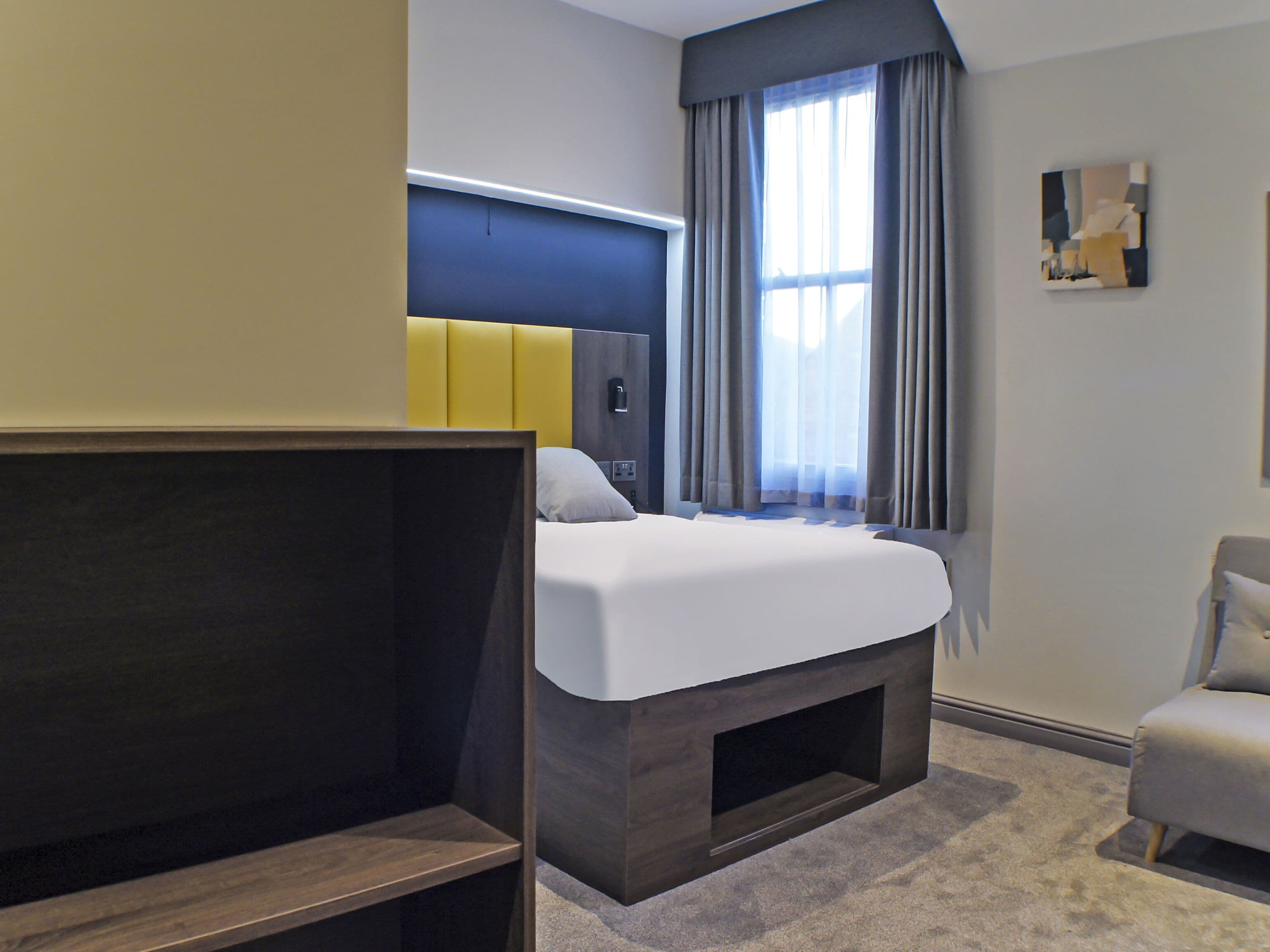 Quarto de hotel moderno com uma cama grande, cabeceira, área de estar e janela com cortinas.