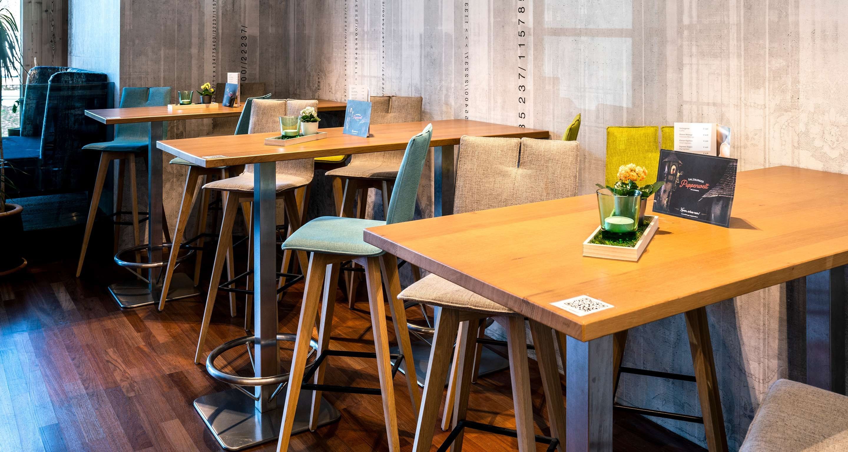 Interior de restaurante com mesas e cadeiras de madeira, algumas com almofadas. Pequenas plantas em vasos decoram as mesas.