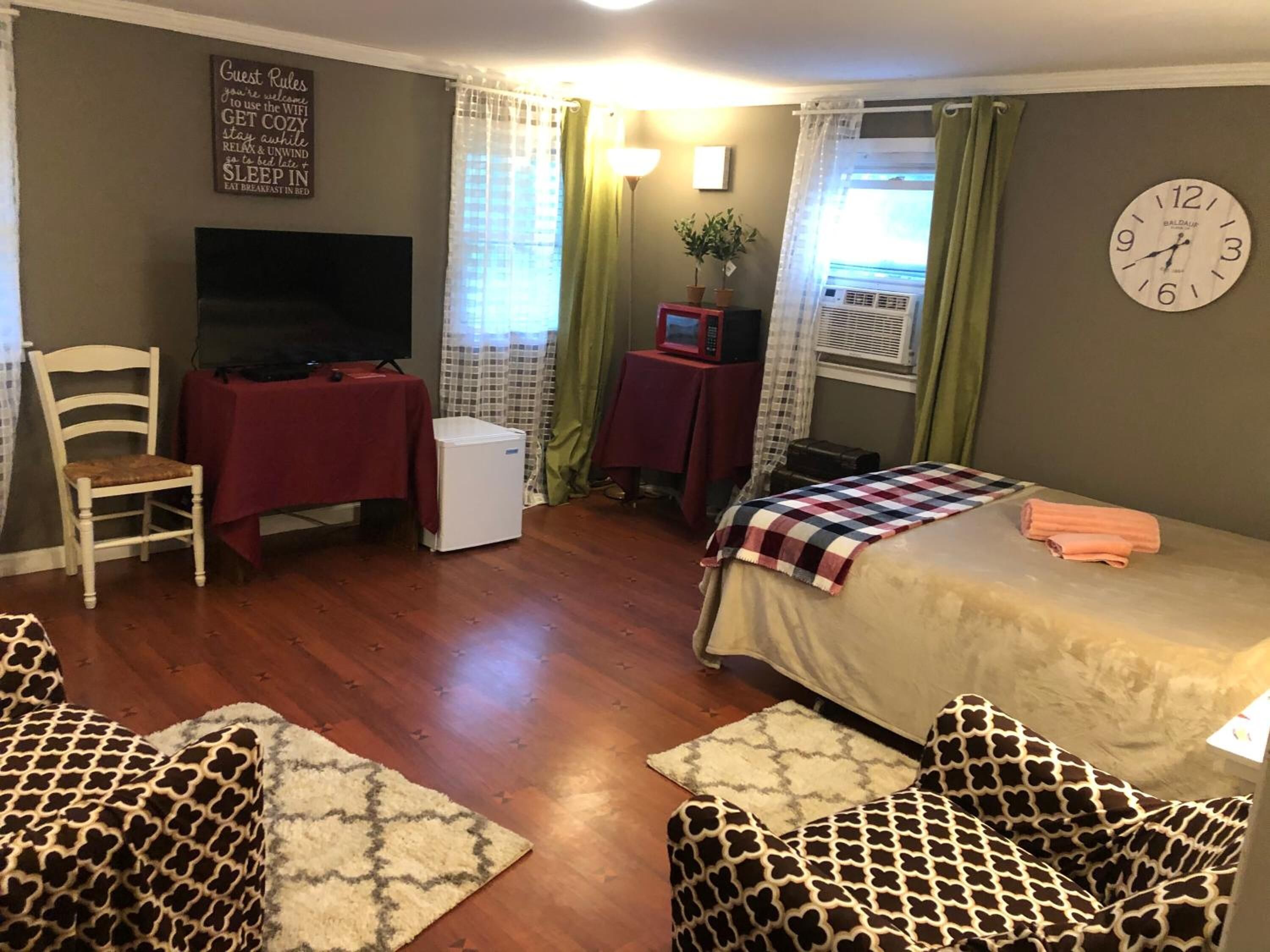 Quarto de hotel aconchegante com cama, televisão, mini-geladeira, escrivaninha com luminária e relógio na parede.