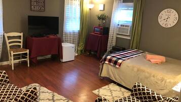 Quarto de hotel aconchegante com cama, televisão, mini-geladeira, escrivaninha com luminária e relógio na parede.