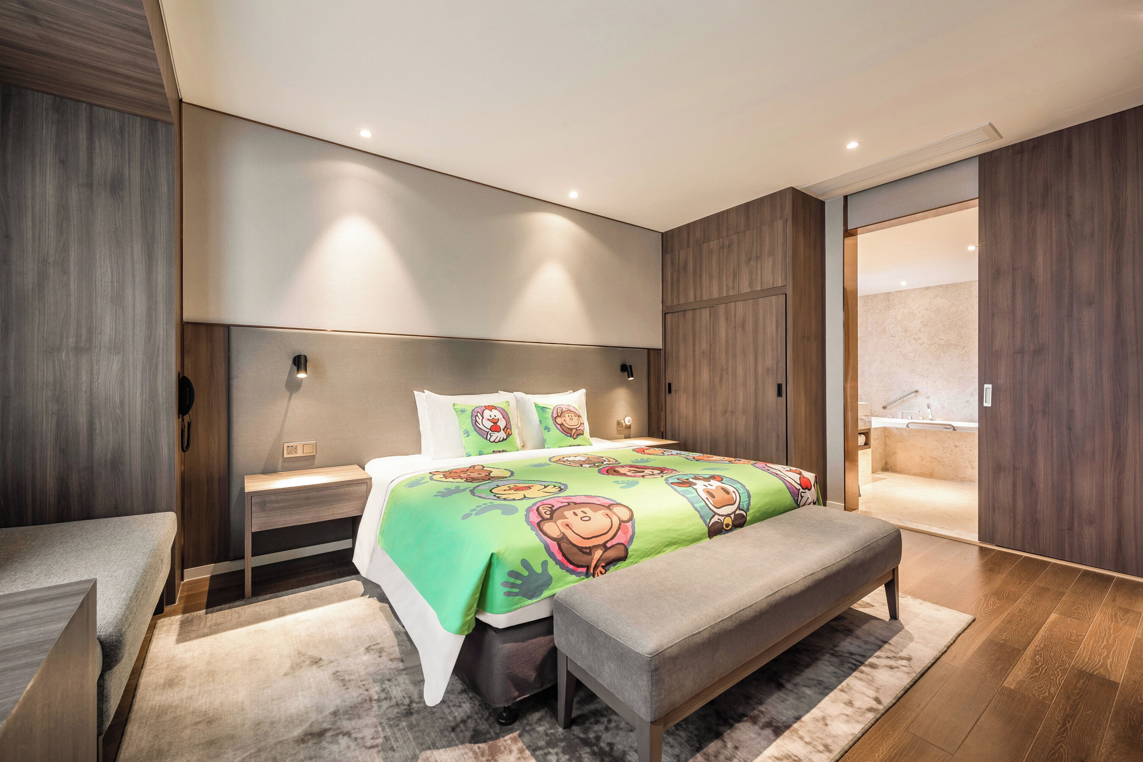 Quarto moderno com uma cama grande, mesas de cabeceira, um banco e um banheiro visível por uma porta aberta.