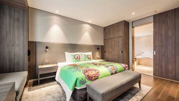 Quarto moderno com uma cama grande, mesas de cabeceira, um banco e um banheiro visível por uma porta aberta.