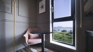 Quarto com vista para um corpo d'água e montanhas, uma cadeira rosa, uma pequena mesa redonda e um mastro dourado.