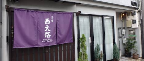 Um edifício com uma faixa roxa que diz "Guest House Nishioji" e um portão marrom. Há plantas em vasos na calçada.