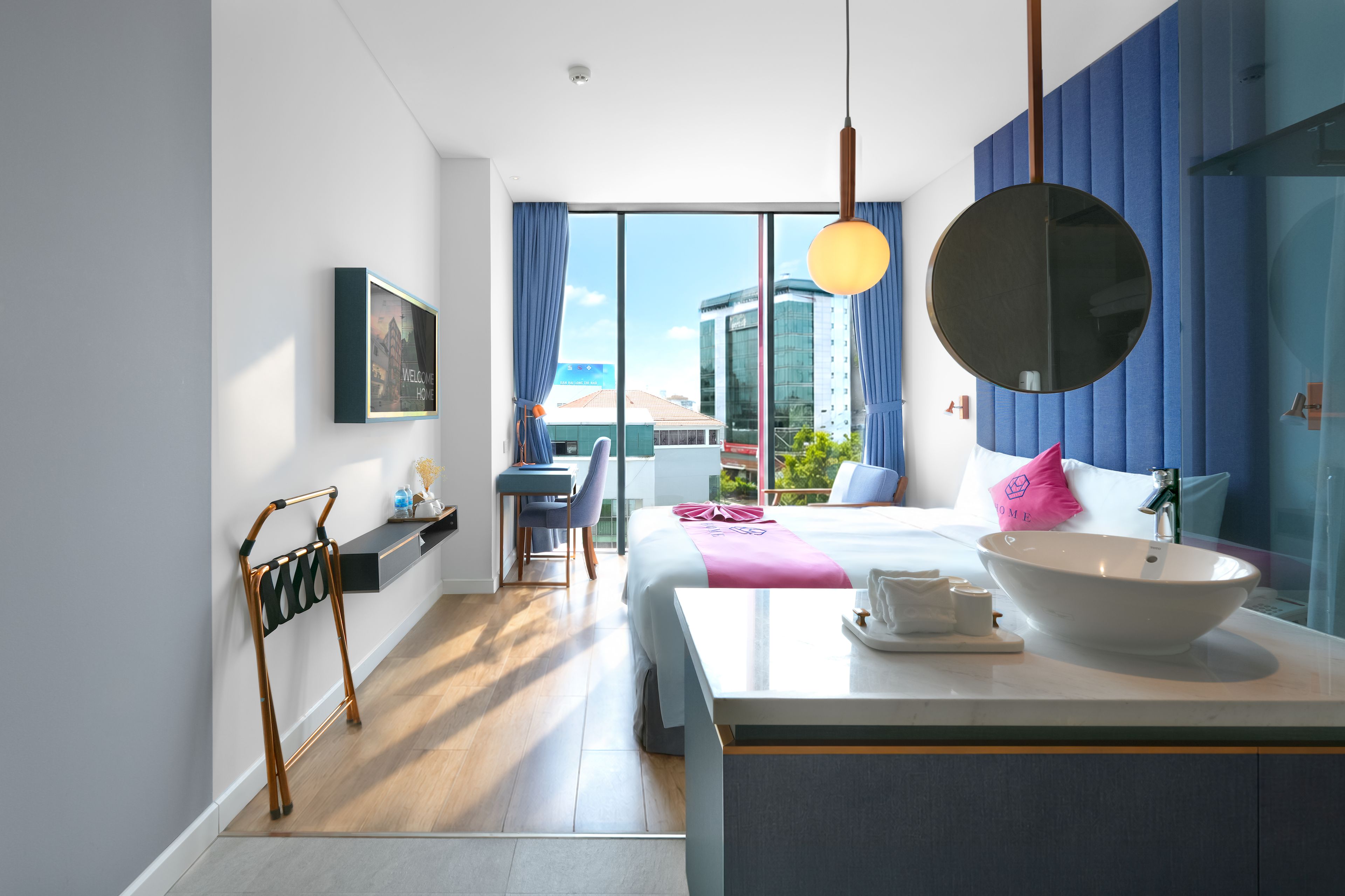 Quarto de hotel moderno com uma cama grande, um espelho redondo, uma mesa de cabeceira com uma pia branca, uma escrivaninha com uma cadeira e vista para a cidade através de amplas janelas.