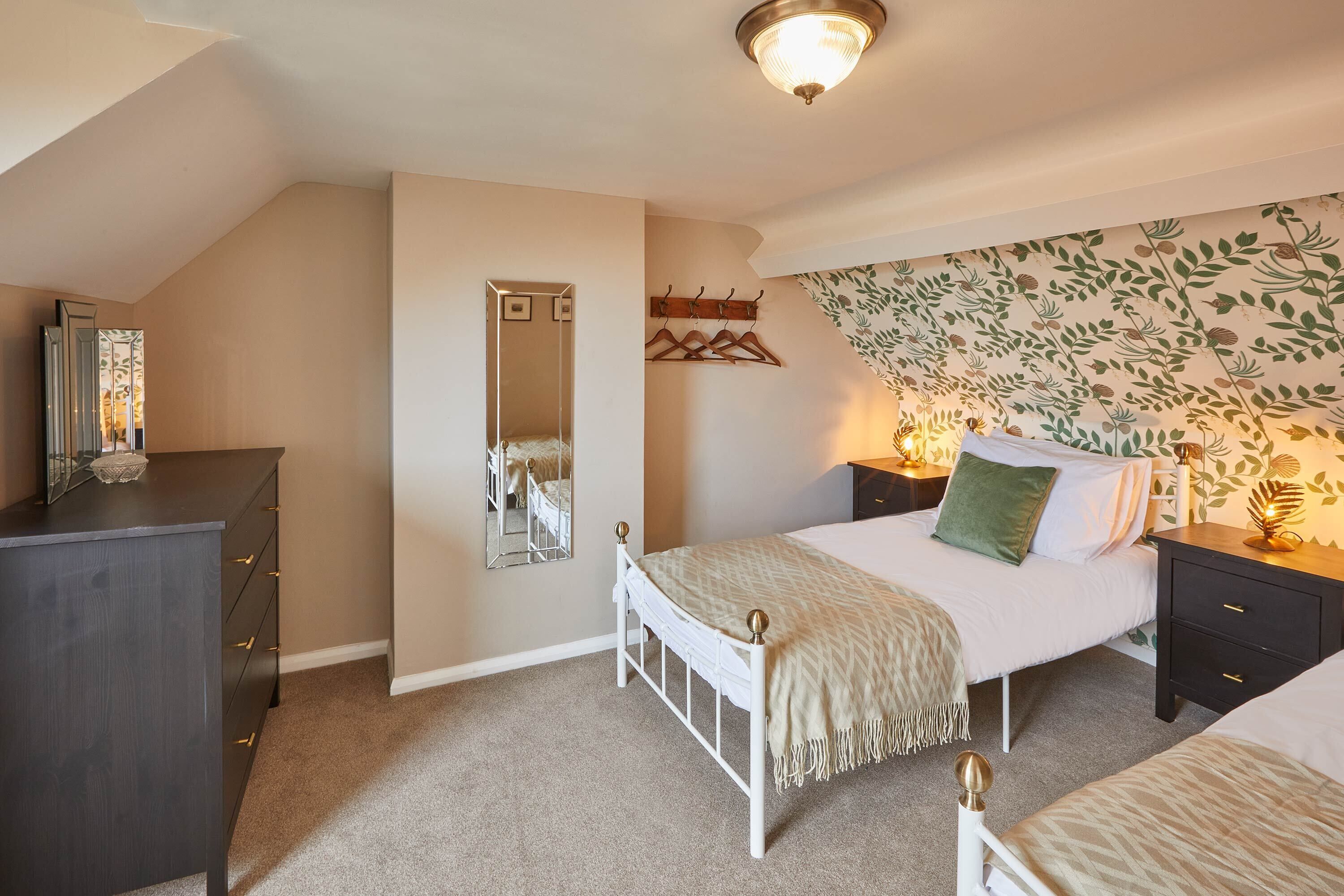 Quarto com cama, mesas de cabeceira, cômoda, espelho e parede com padrão de folhas.