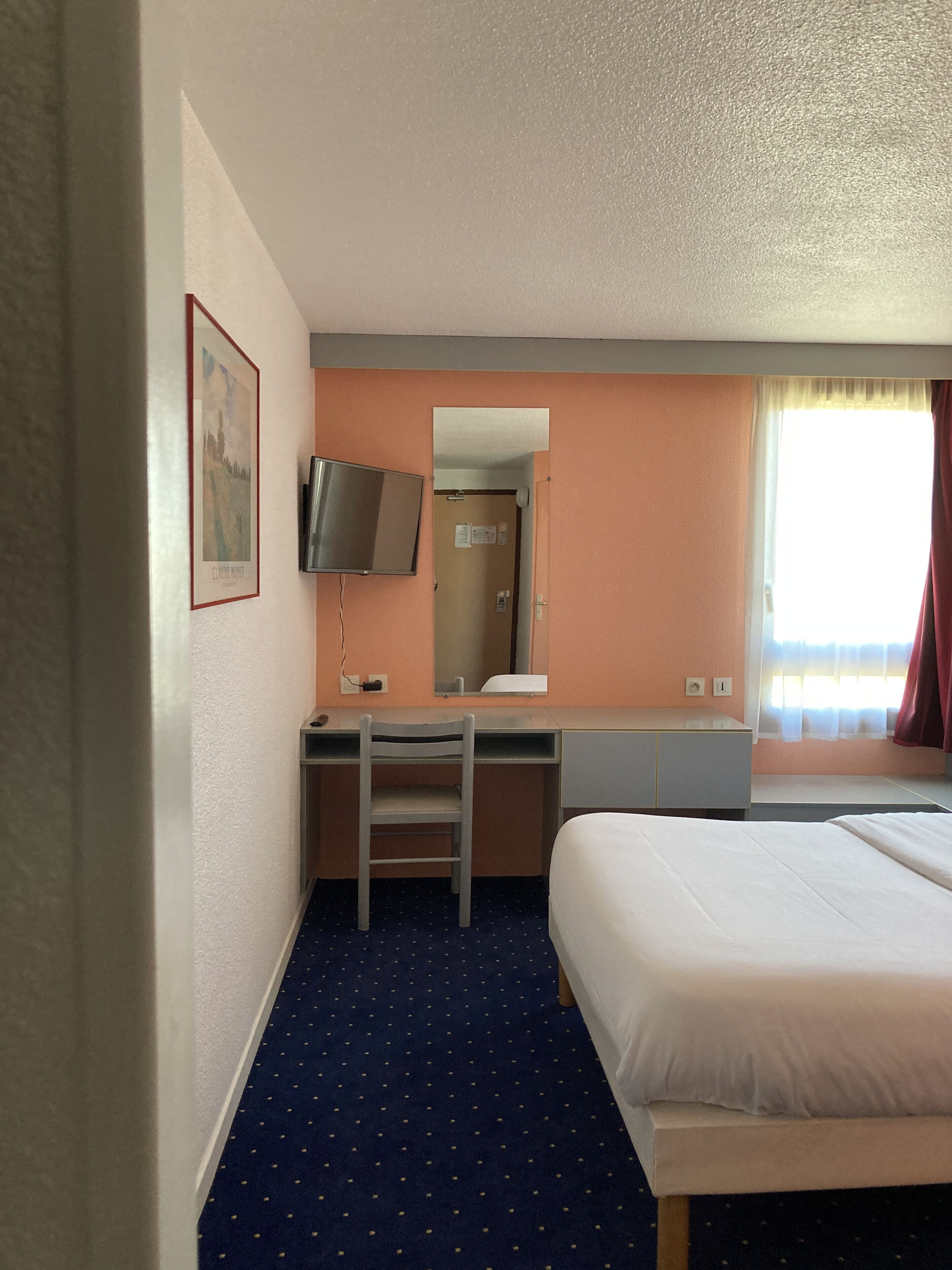 Quarto de hotel com cama, escrivaninha, cadeira, TV fixada na parede e janela com cortinas.