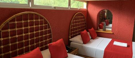 Quarto de hotel com duas camas, roupa de cama vermelha e branca, uma janela grande com vista para vegetação e uma parede vermelha texturizada.