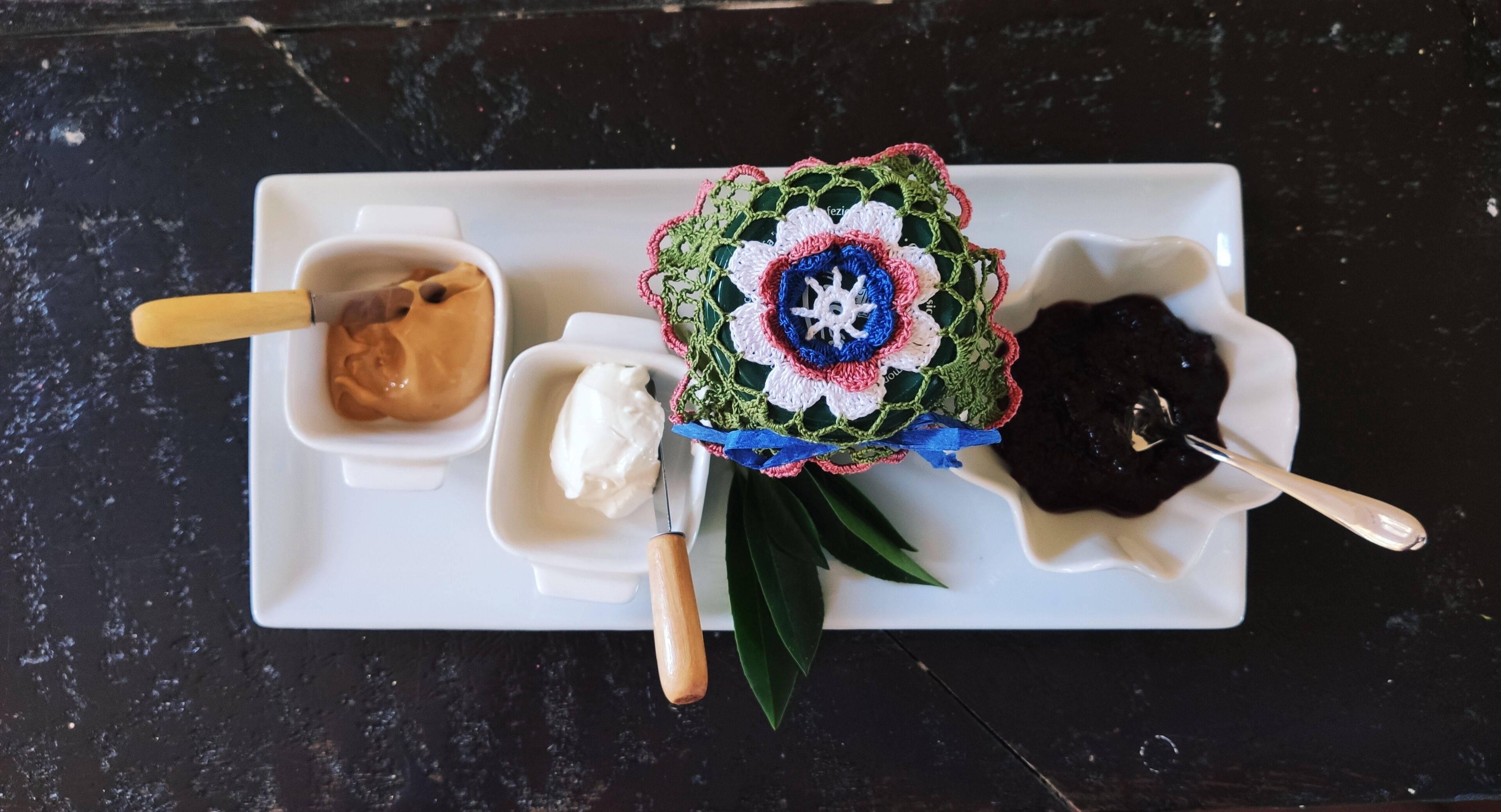 Uma bandeja retangular branca com três pequenas tigelas contendo diferentes alimentos, uma colher de cabo de madeira e um centro de mesa de crochê com padrão floral em azul, branco e vermelho.