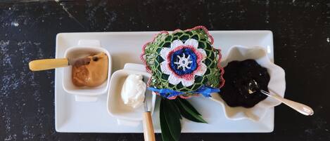 Uma bandeja retangular branca com três pequenas tigelas contendo diferentes alimentos, uma colher de cabo de madeira e um centro de mesa de crochê com padrão floral em azul, branco e vermelho.