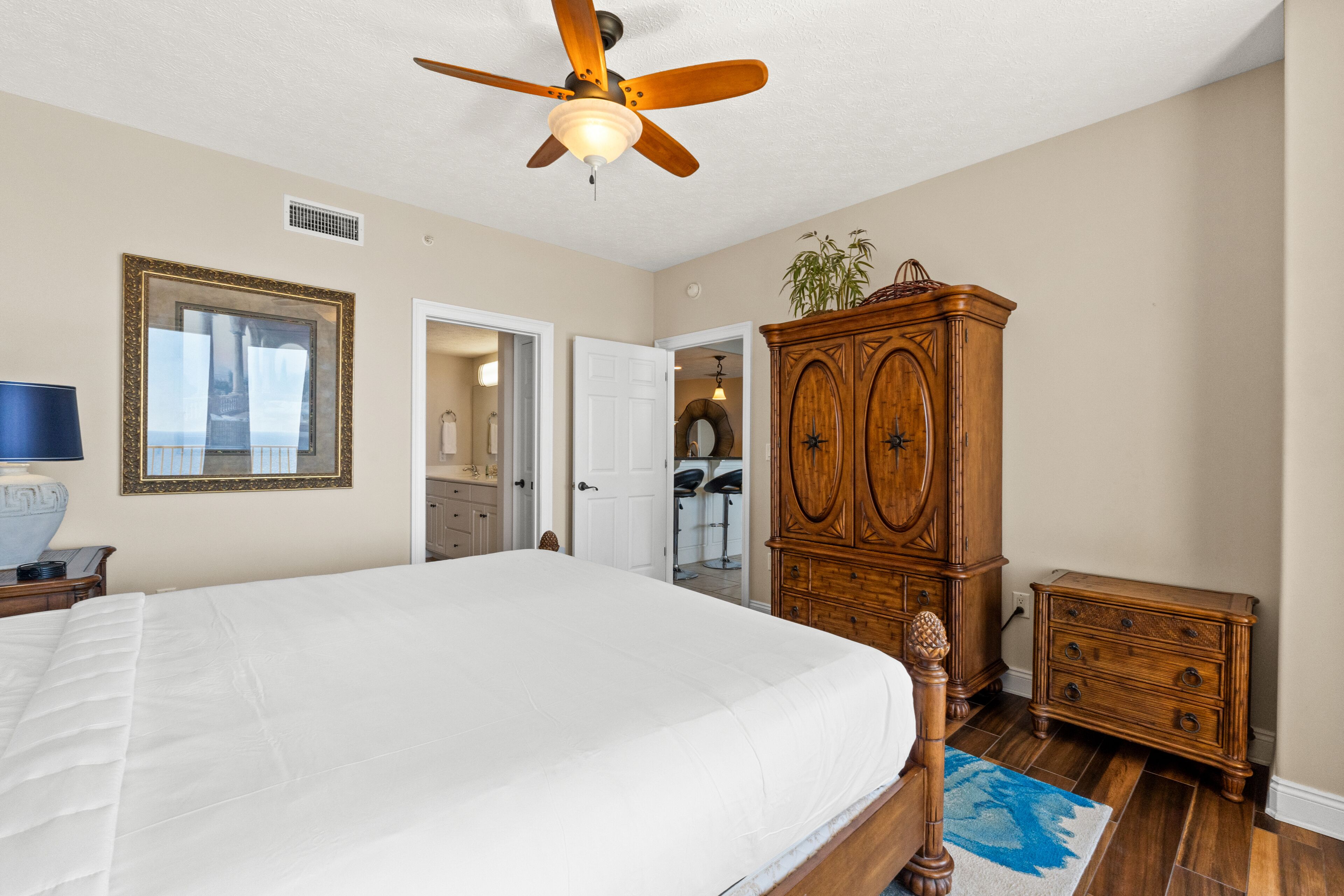 Quarto com cama grande, cómoda de madeira, ventoinha no teto e vista para o banheiro.